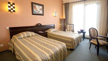 Quarto de hotel com duas camas, uma escrivaninha, uma cadeira e uma janela grande com cortinas.