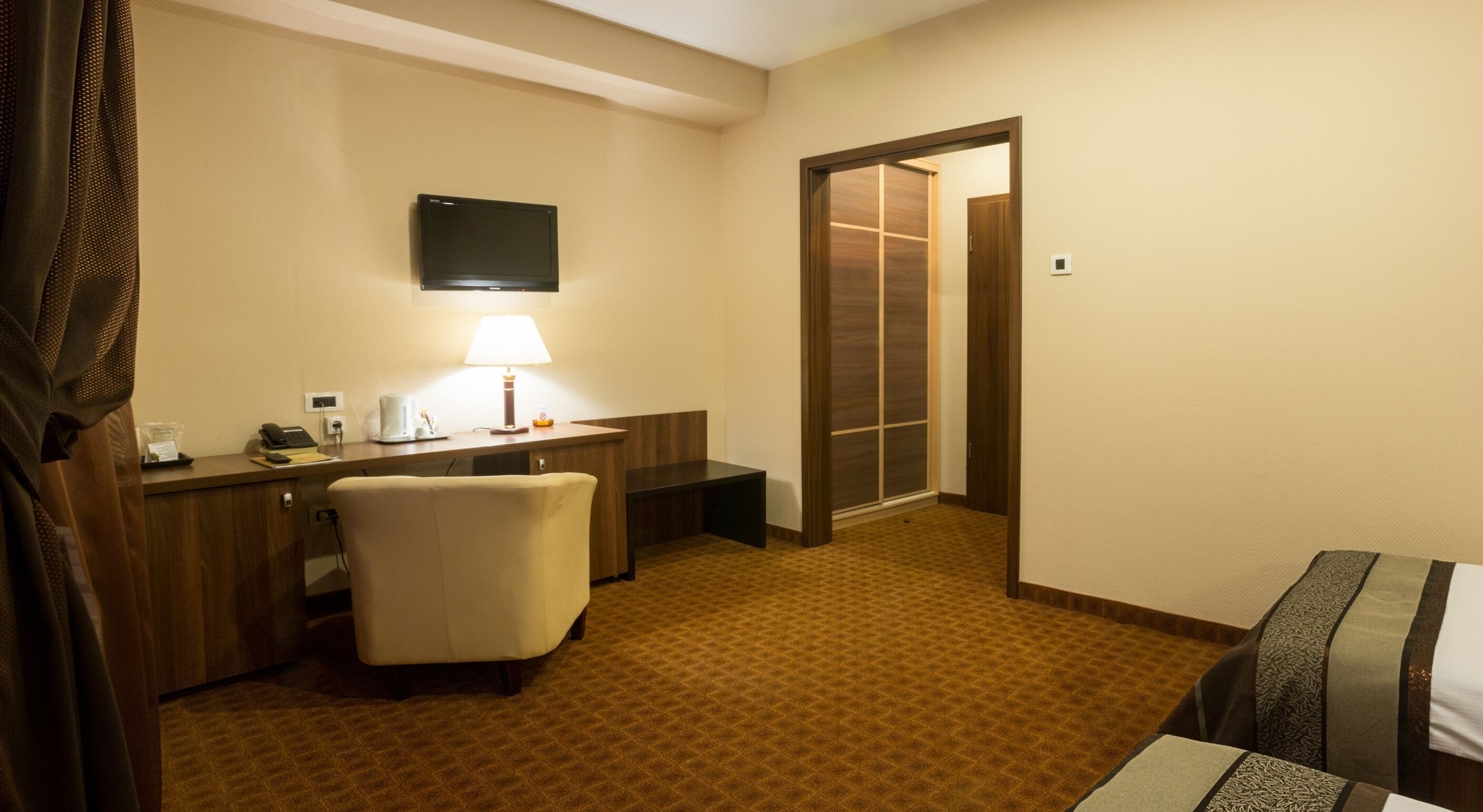 Quarto de hotel com escrivaninha, cadeira, cama e televisão fixada na parede.