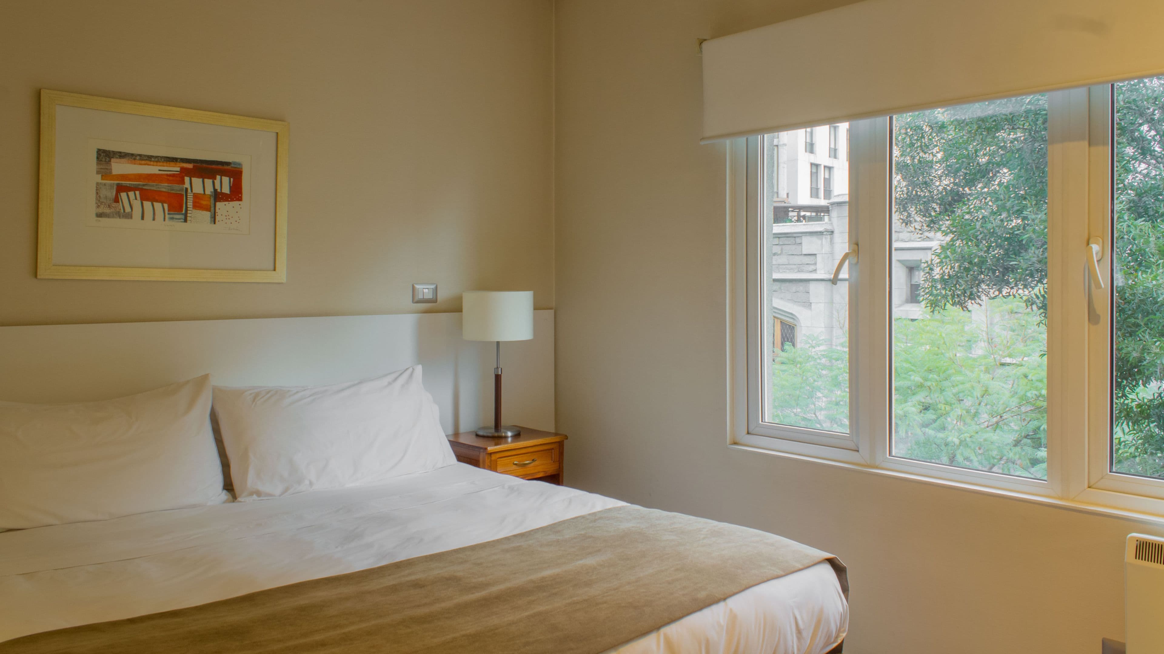 Cama bem arrumada com lençóis brancos e manta marrom, mesa de cabeceira com abajur e janela com vista para área verde e edifícios.