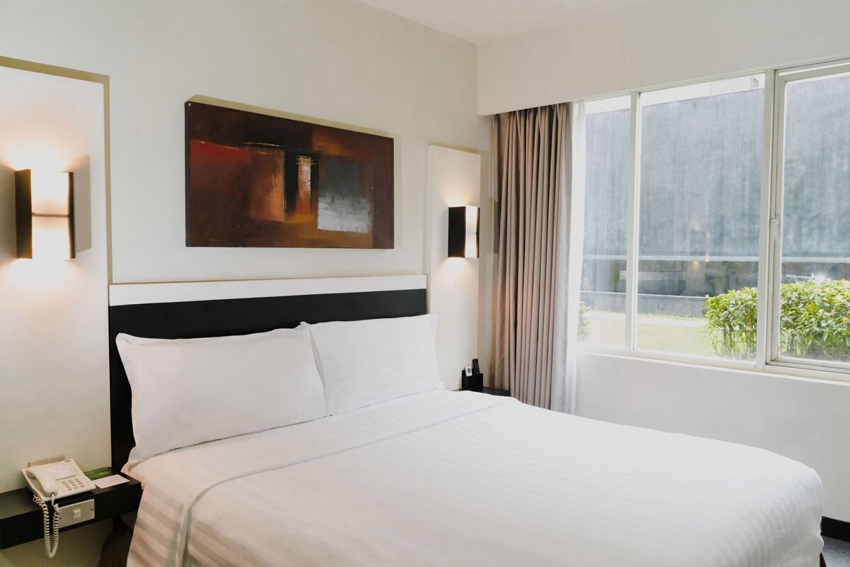 Quarto de hotel com uma cama grande, um quadro na parede, uma mesa de cabeceira com telefone e uma janela com cortinas.