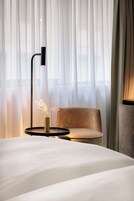 Quarto de hotel com cama, uma cadeira, mesa com um vaso de flores e abajur.