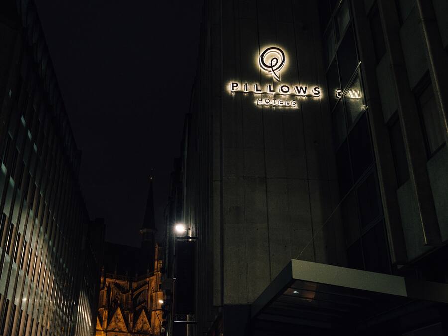 Exterior do Hotel Pillow's à noite, com fachada moderna e letreiros iluminados.