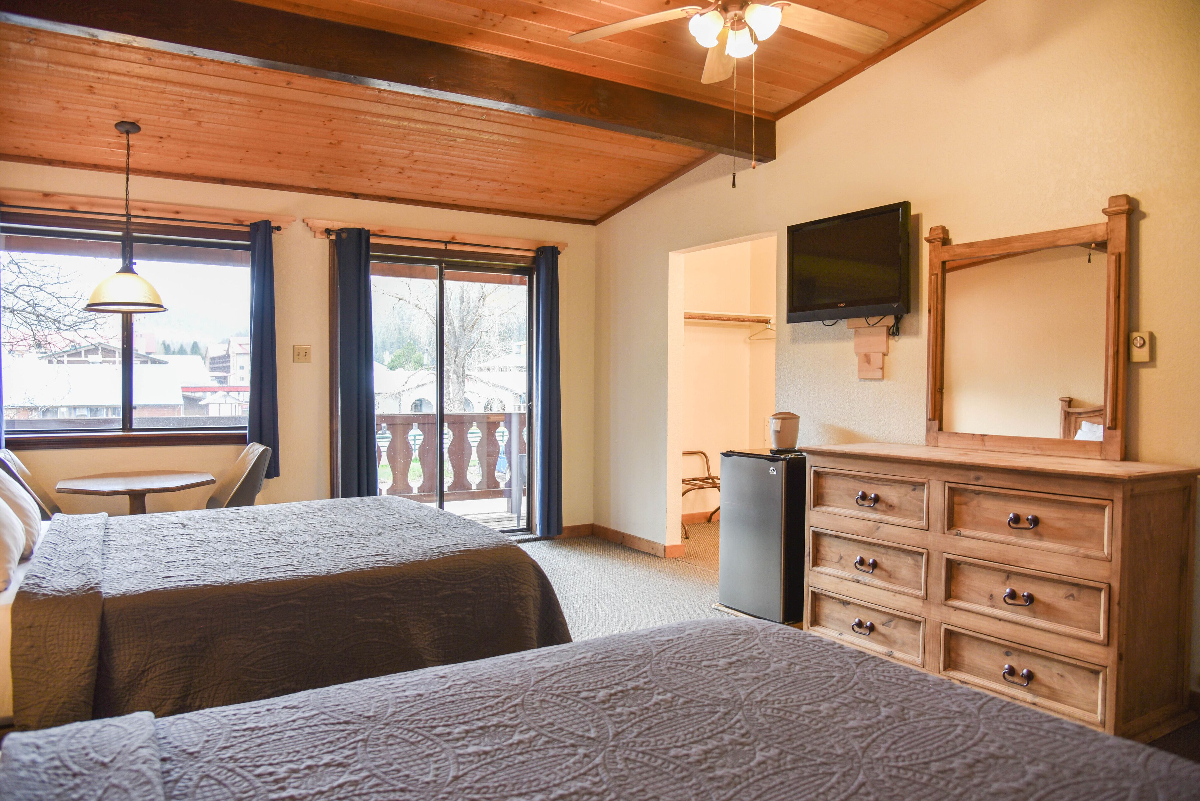 Quarto de hotel com duas camas, cômoda de madeira, televisão e varanda com mesa e cadeiras.