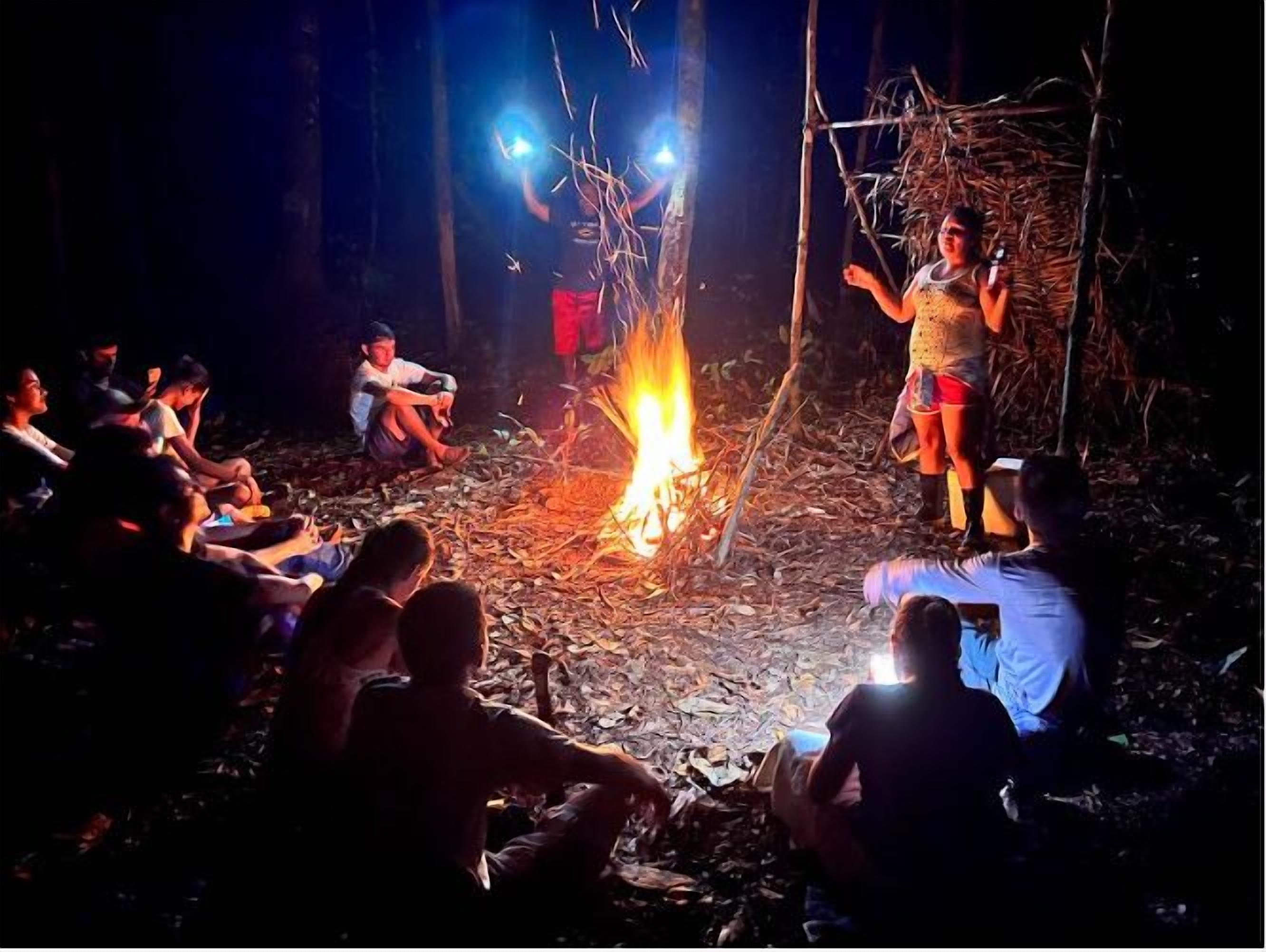 Um grupo de pessoas sentadas ao redor de uma fogueira em uma área arborizada.