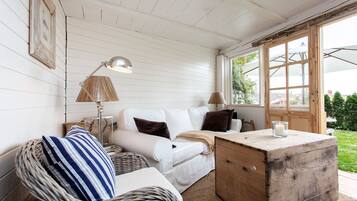 Um quarto aconchegante com teto de madeira branca, um sofá branco com almofadas marrons, uma cadeira de vime, um baú de madeira e um abajur.