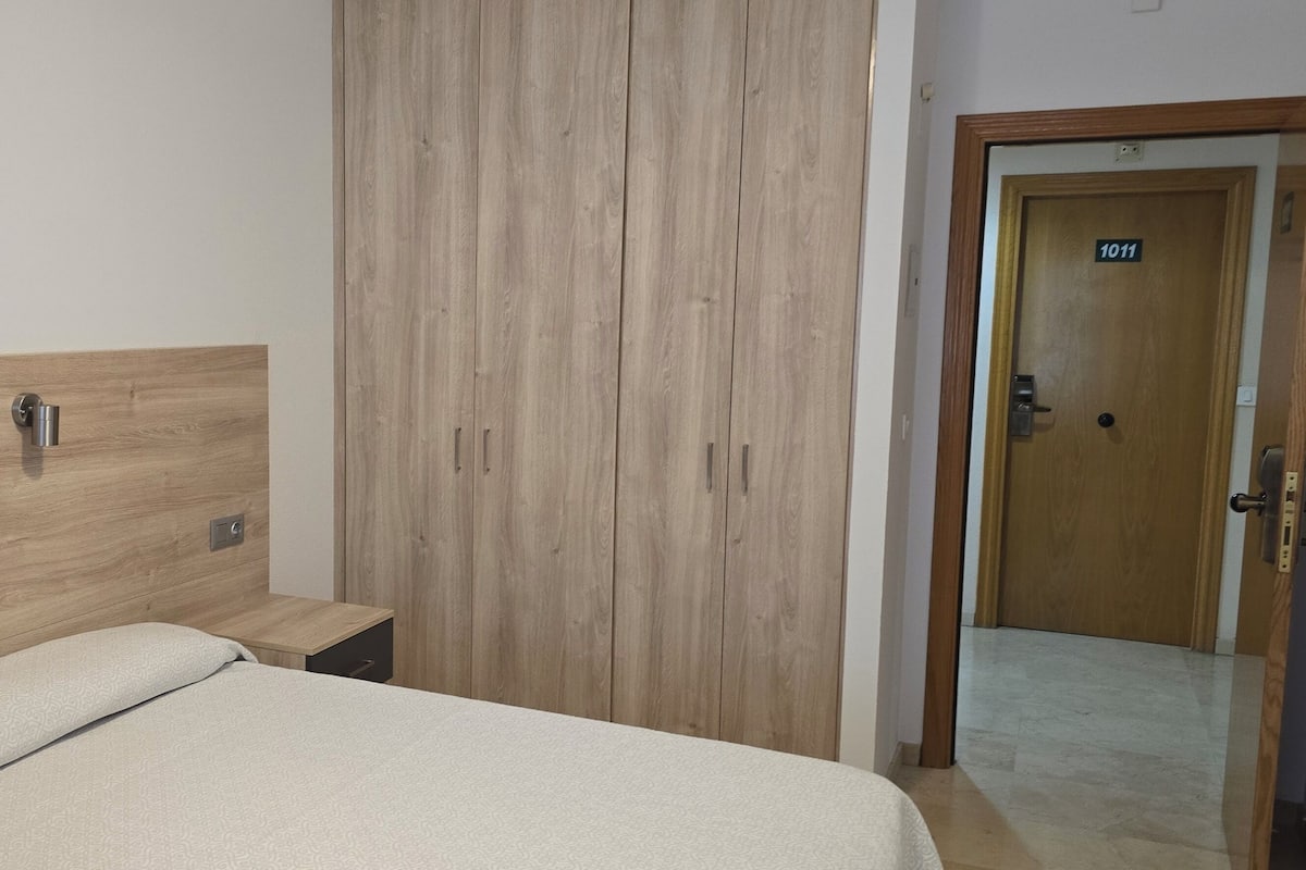 Quarto com cabeceira de madeira, cama com colchão branco, guarda-roupa de madeira e uma porta com a inscrição 1011.