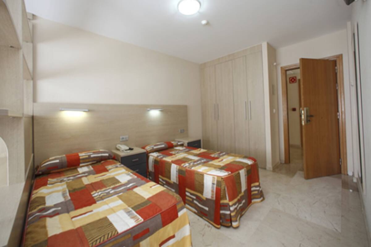 Quarto com duas camas, um guarda-roupa, uma mesa de cabeceira e uma porta que dá para outro espaço.