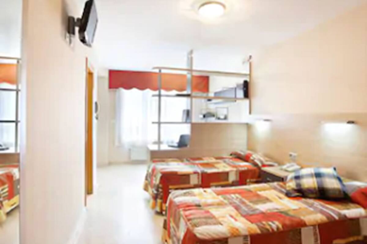Quarto de estudante com duas camas, uma televisão, uma estante e uma janela.
