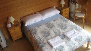 Um quarto de madeira com uma cama, duas mesas de cabeceira, uma cadeira e um aquecedor fixado na parede.