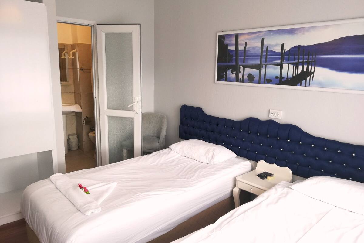 Quarto de hotel com duas camas, banheiro e pintura de uma paisagem à beira de um lago.