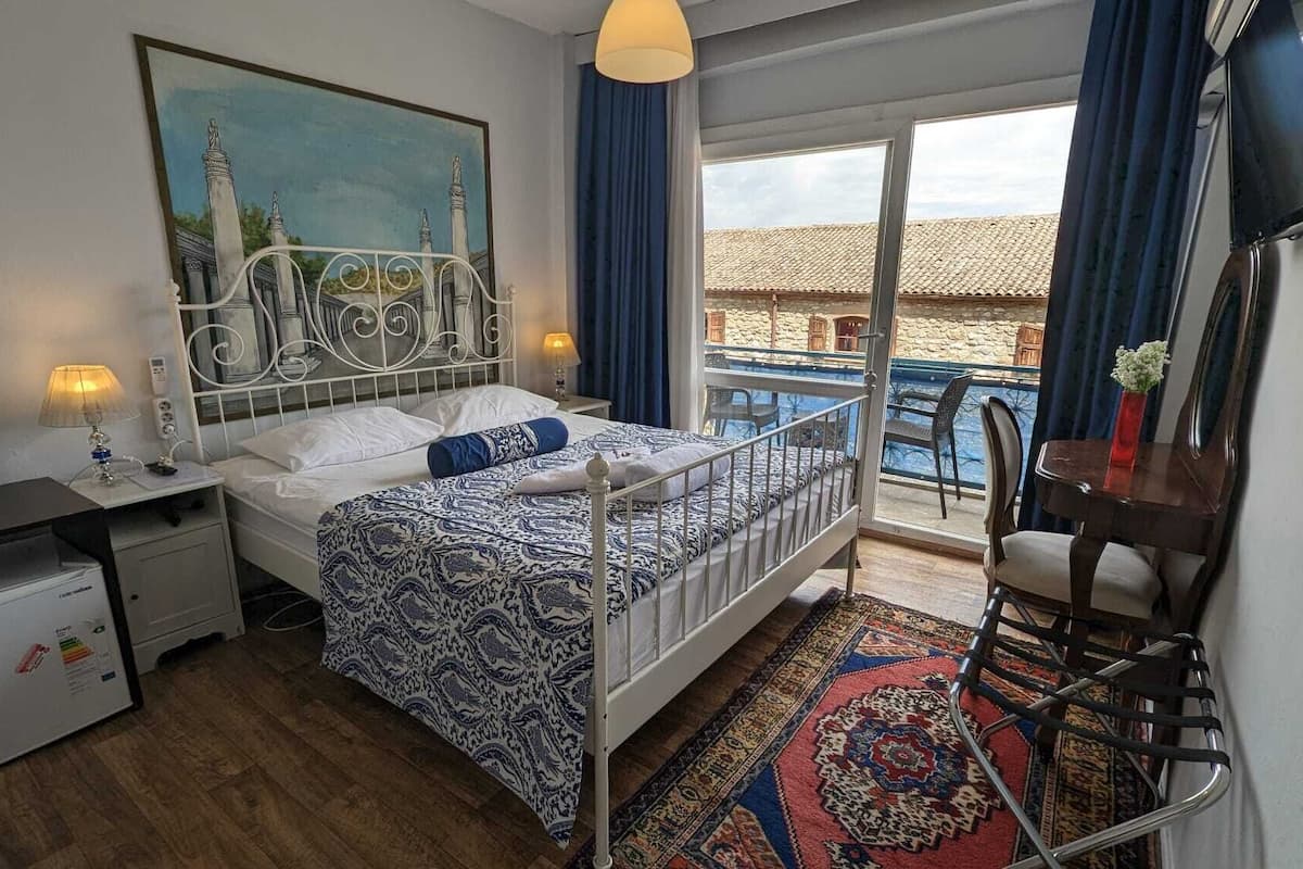 Quarto de hotel com cama, escrivaninha, cadeira, mini-geladeira, pintura na parede e varanda com vista para edifícios.