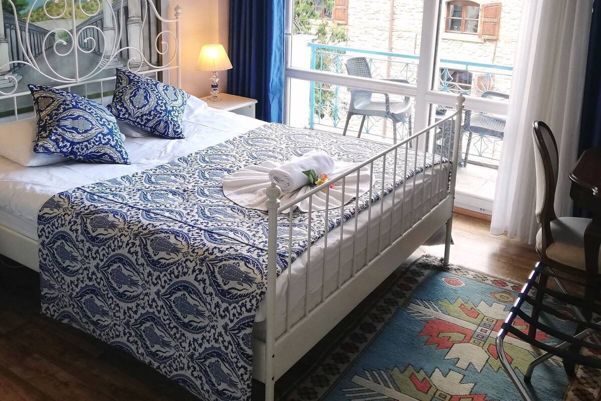 Quarto com cama, um quadro, janela com cortinas e vista para prédios lá fora.