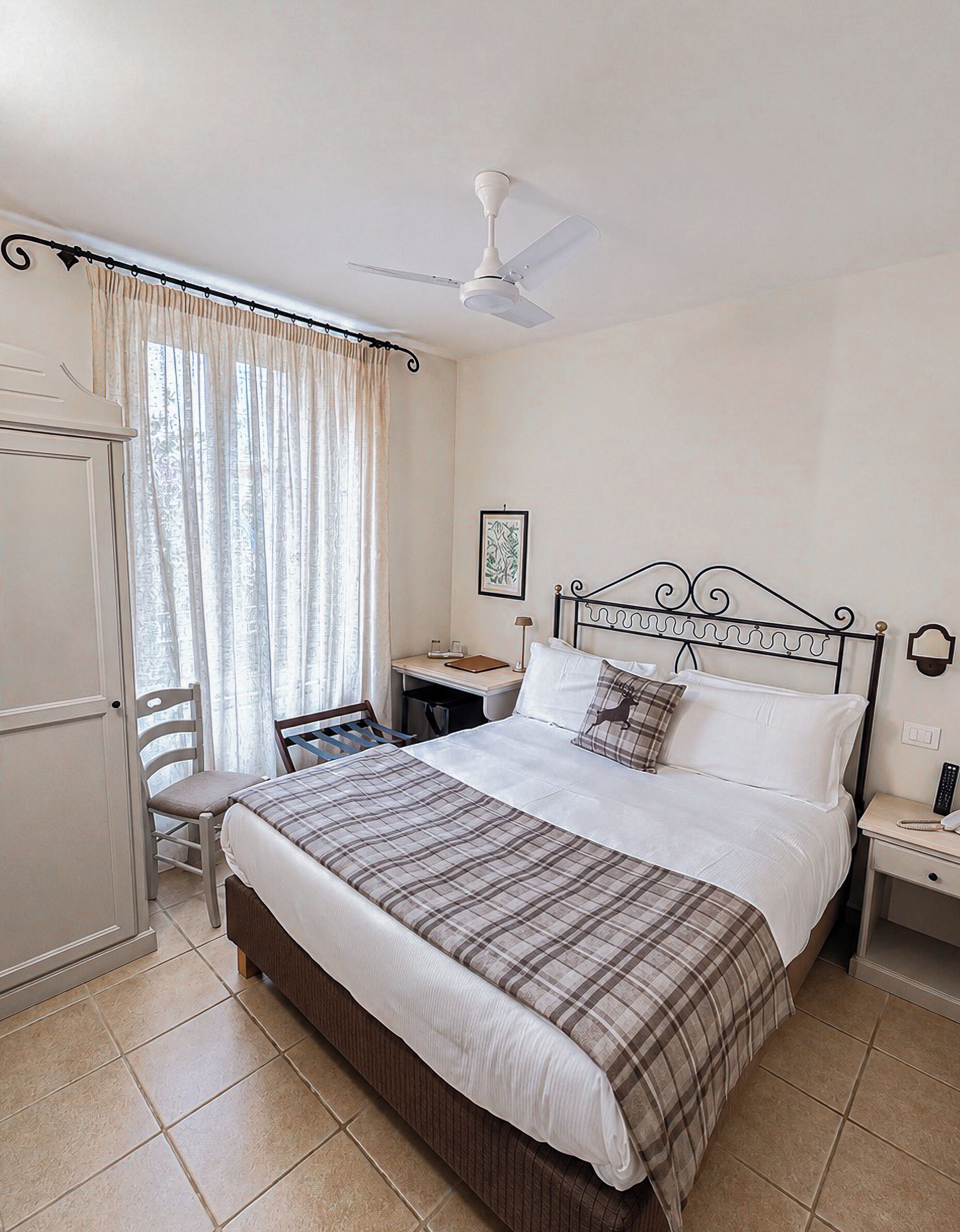 Quarto com cama, escrivaninha, cadeira, guarda-roupa, ventoinha no teto e janela com cortinas.
