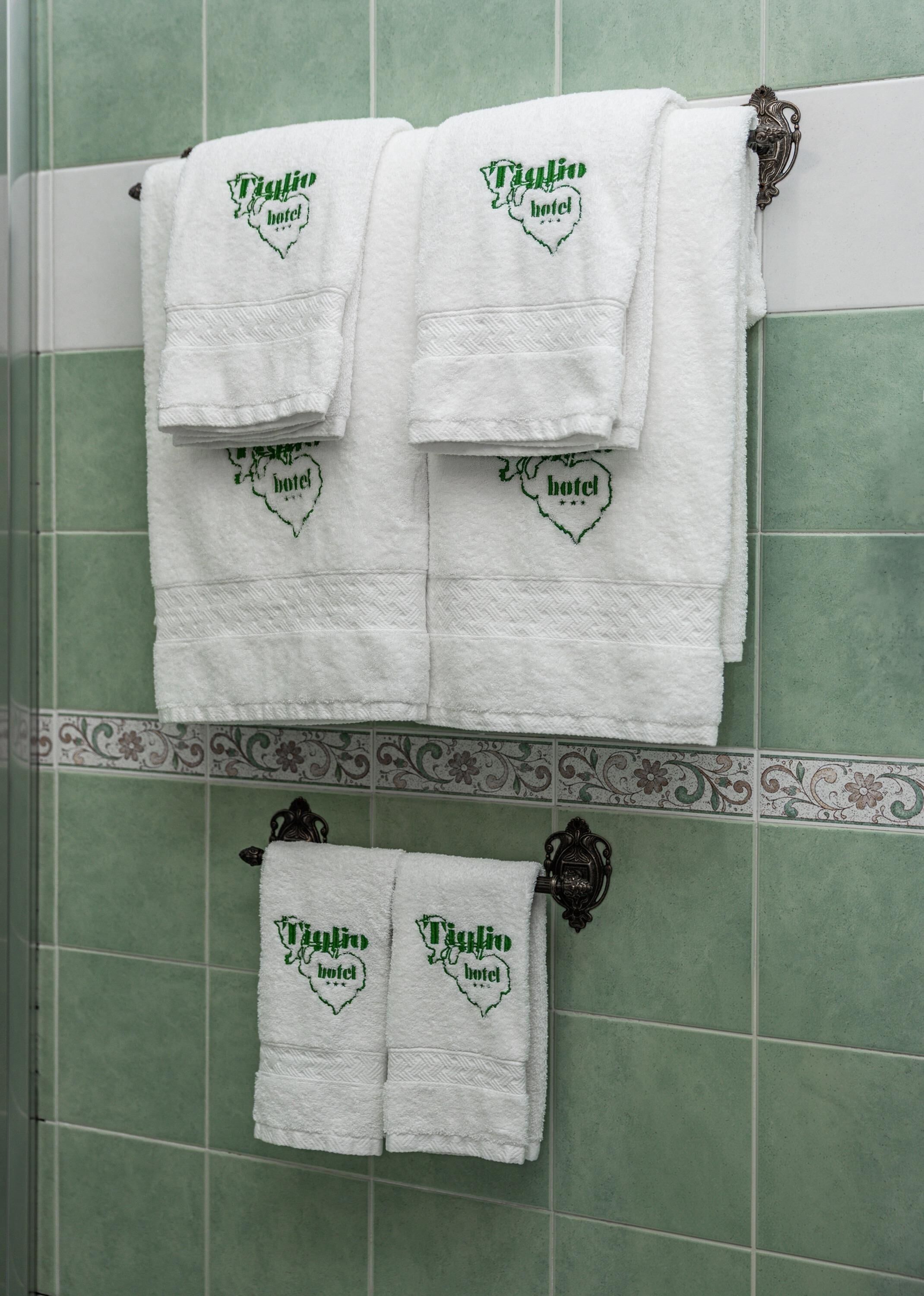 Casa de banho com azulejos verdes e duas toalhas brancas penduradas num suporte, cada uma com bordado 'Taglio hotel'.