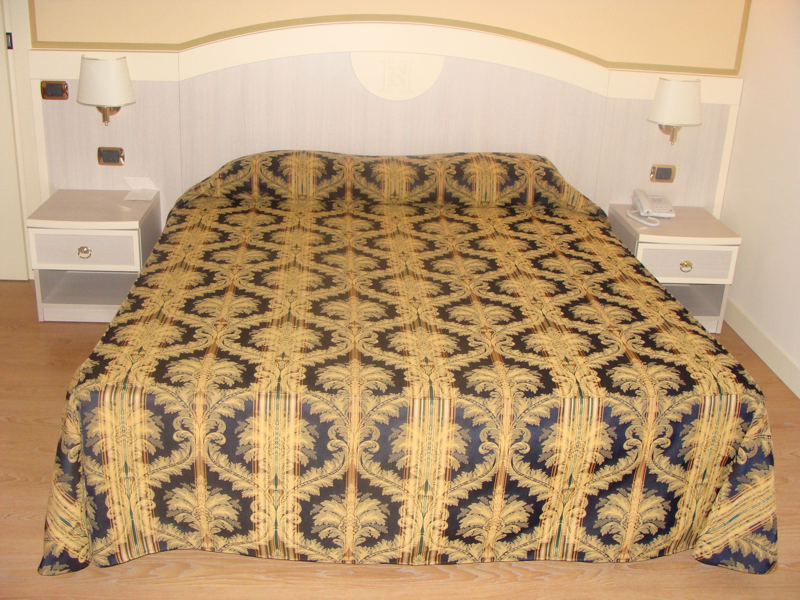Uma cama com colcha estampada, duas mesas de cabeceira com abajures e um criado-mudo com telefone.