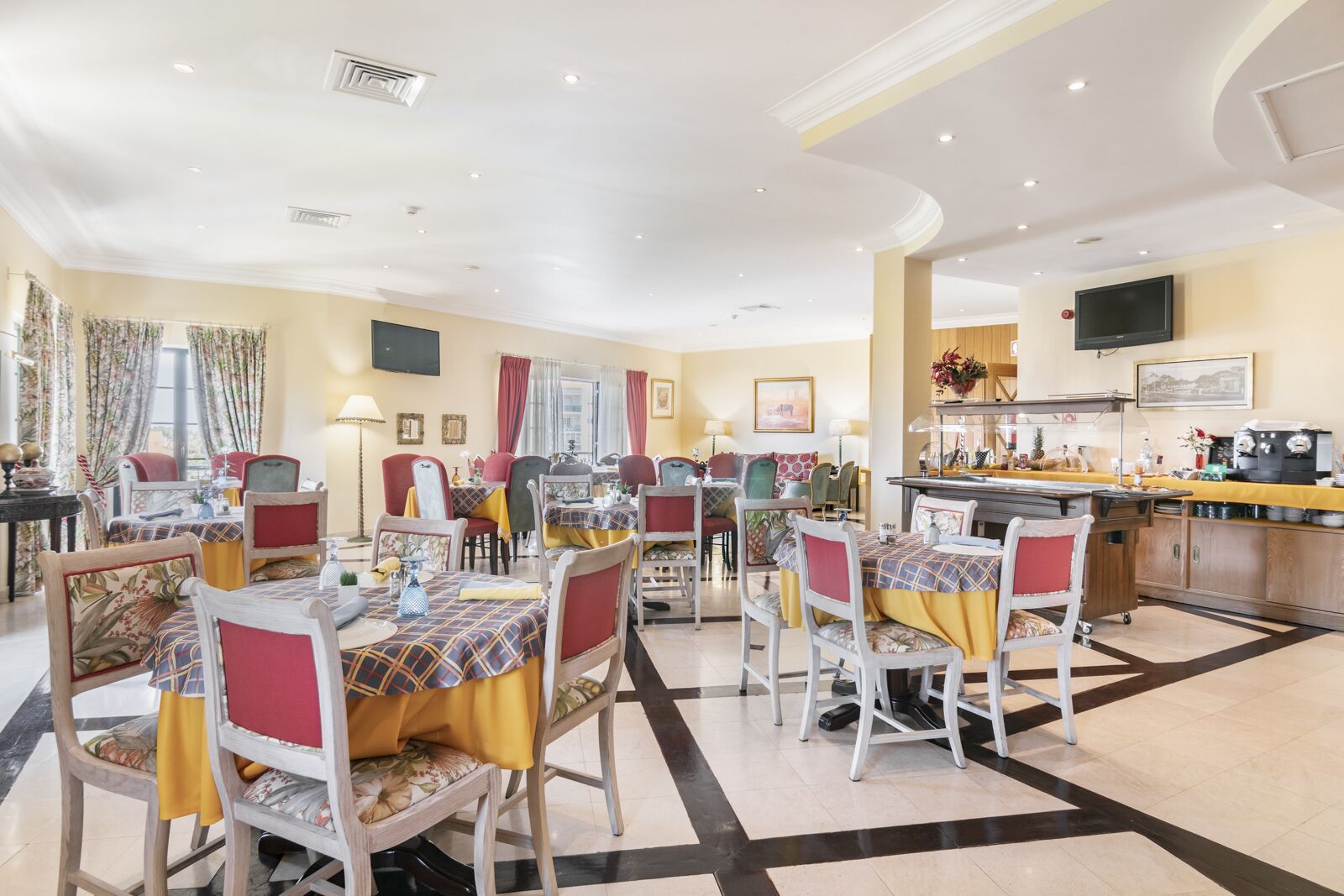 Uma sala de jantar com várias mesas e cadeiras, área de cozinha com máquina de café e balcão, e uma televisão fixada na parede.