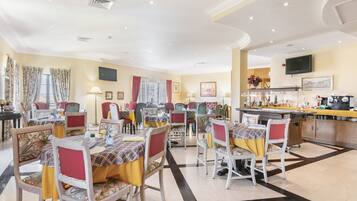 Uma sala de jantar com várias mesas e cadeiras, área de cozinha com máquina de café e balcão, e uma televisão fixada na parede.