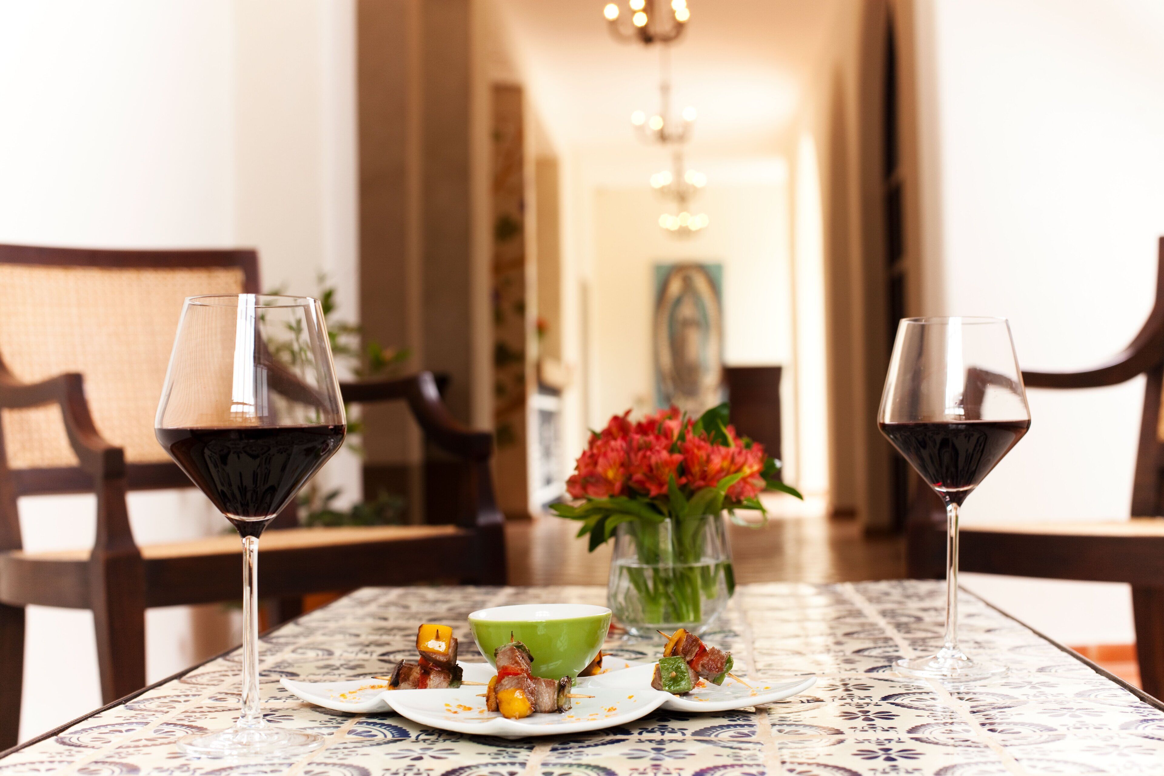 Uma mesa de jantar com duas taças de vinho, uma tigela verde e um vaso de flores vermelhas.