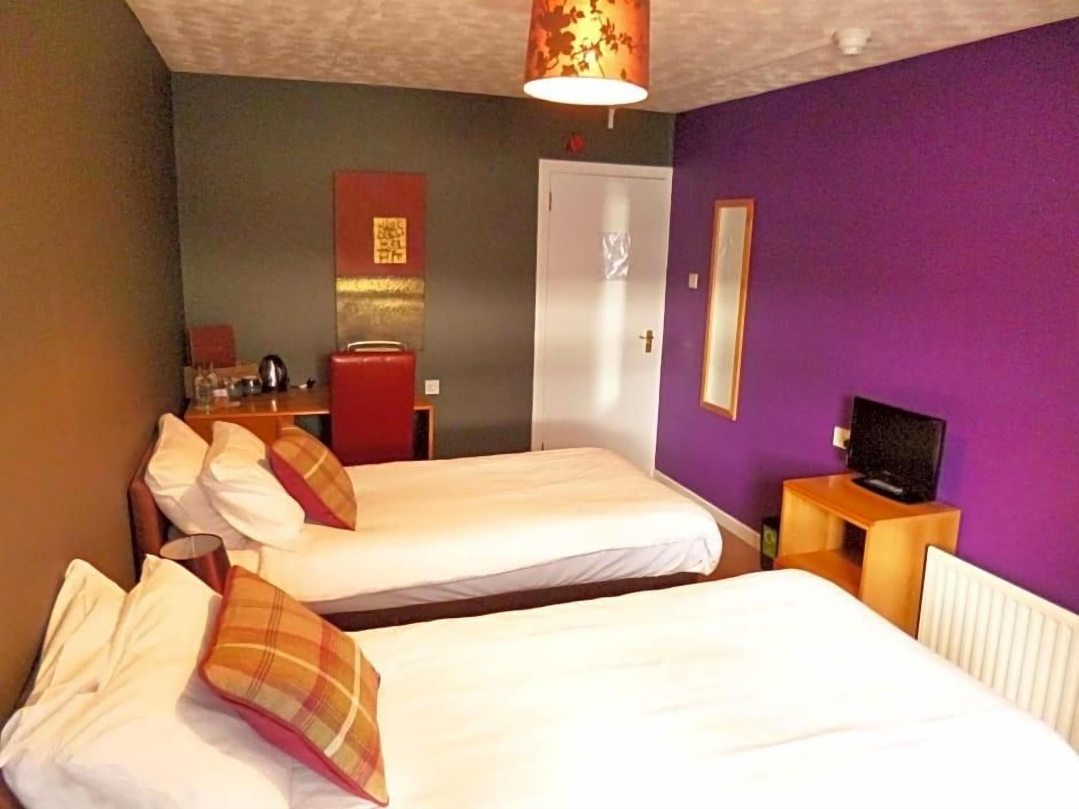 Quarto de hotel com duas camas, uma escrivaninha com computador, uma cadeira vermelha e uma porta.