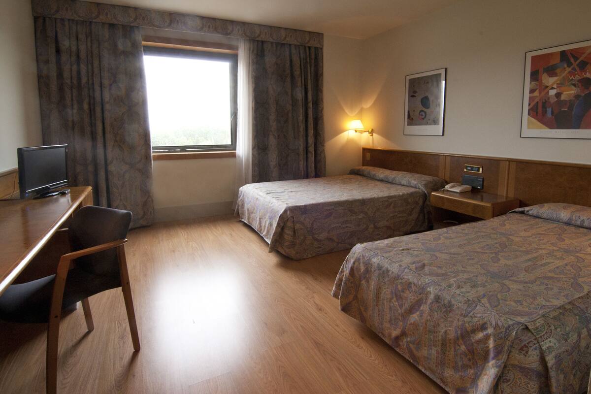 Quarto de hotel com duas camas, uma escrivaninha, uma cadeira, janela com cortinas, luminária de parede e duas fotografias emolduradas.
