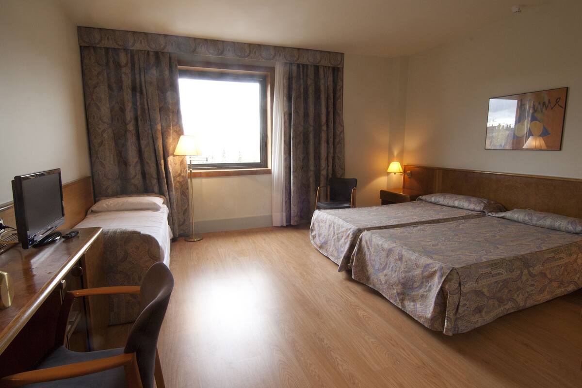 Quarto de hotel com cama, duas mesas de cabeceira, escrivaninha com cadeira, televisão e janela com cortinas.