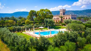Piscina com espreguiçadeiras e guarda-sóis, rodeada por vegetação e um edifício semelhante a um castelo ao fundo.