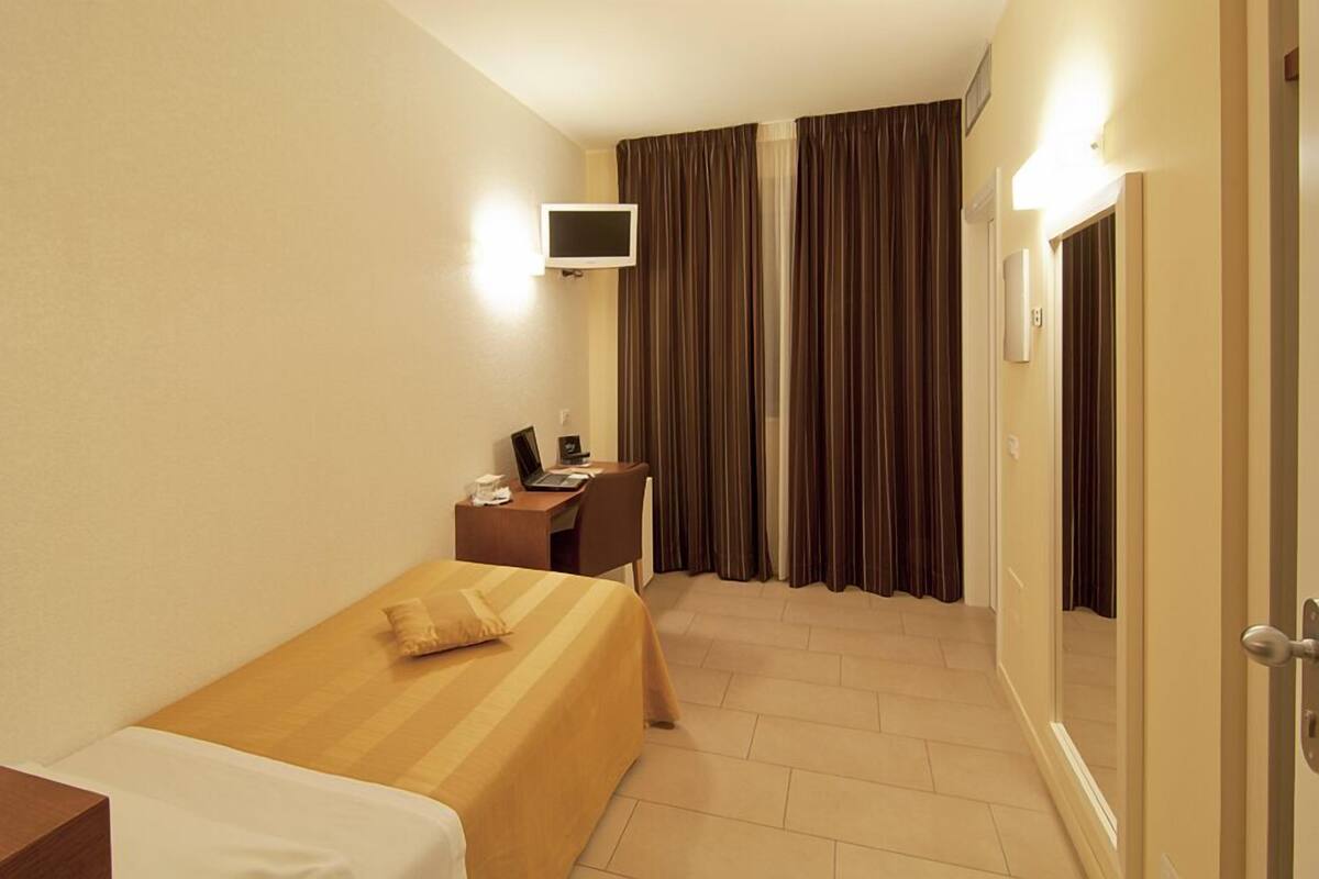 Quarto de hotel com uma cama, uma escrivaninha com computador, televisão e cortinas.