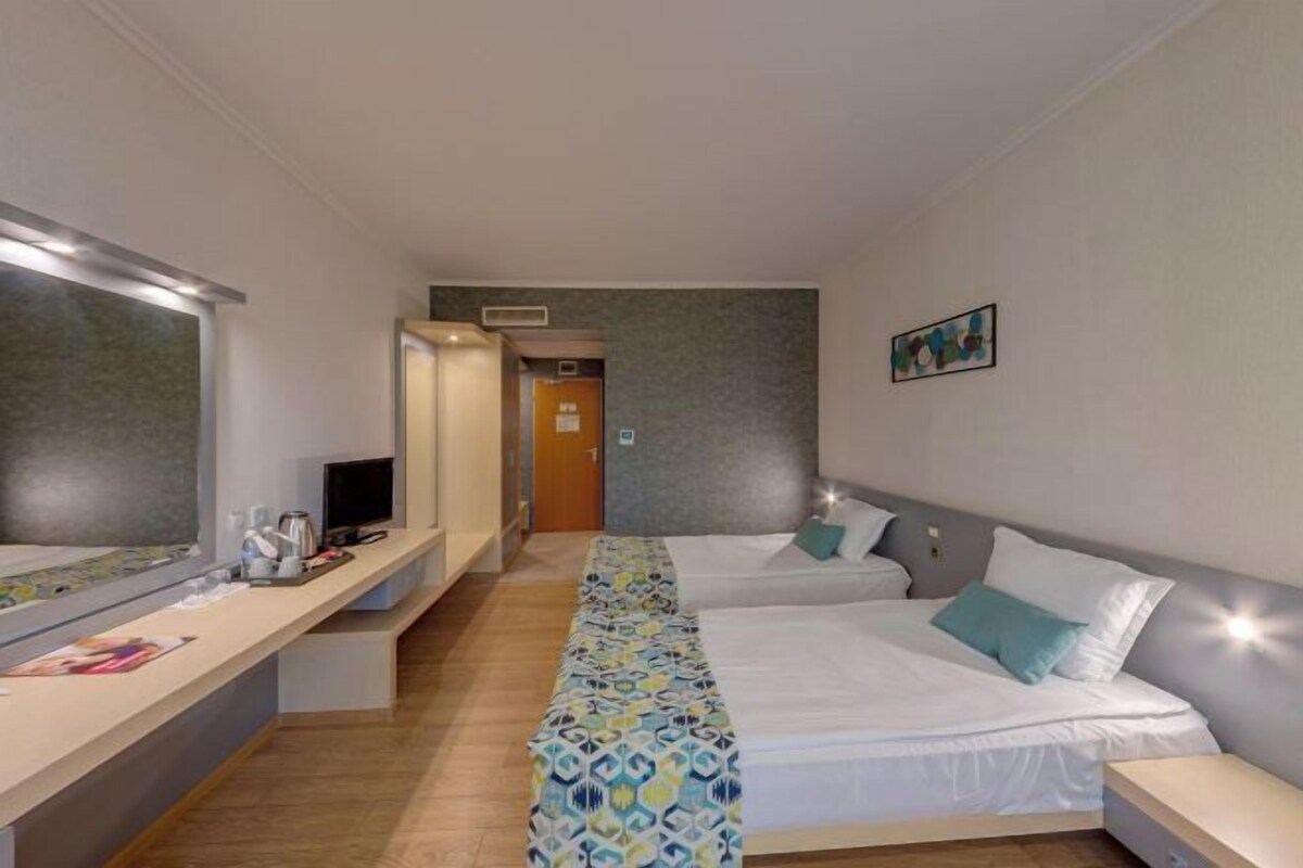 Quarto de hotel com uma cama, mesa de cabeceira e um espelho. Há uma porta que dá para outro quarto.