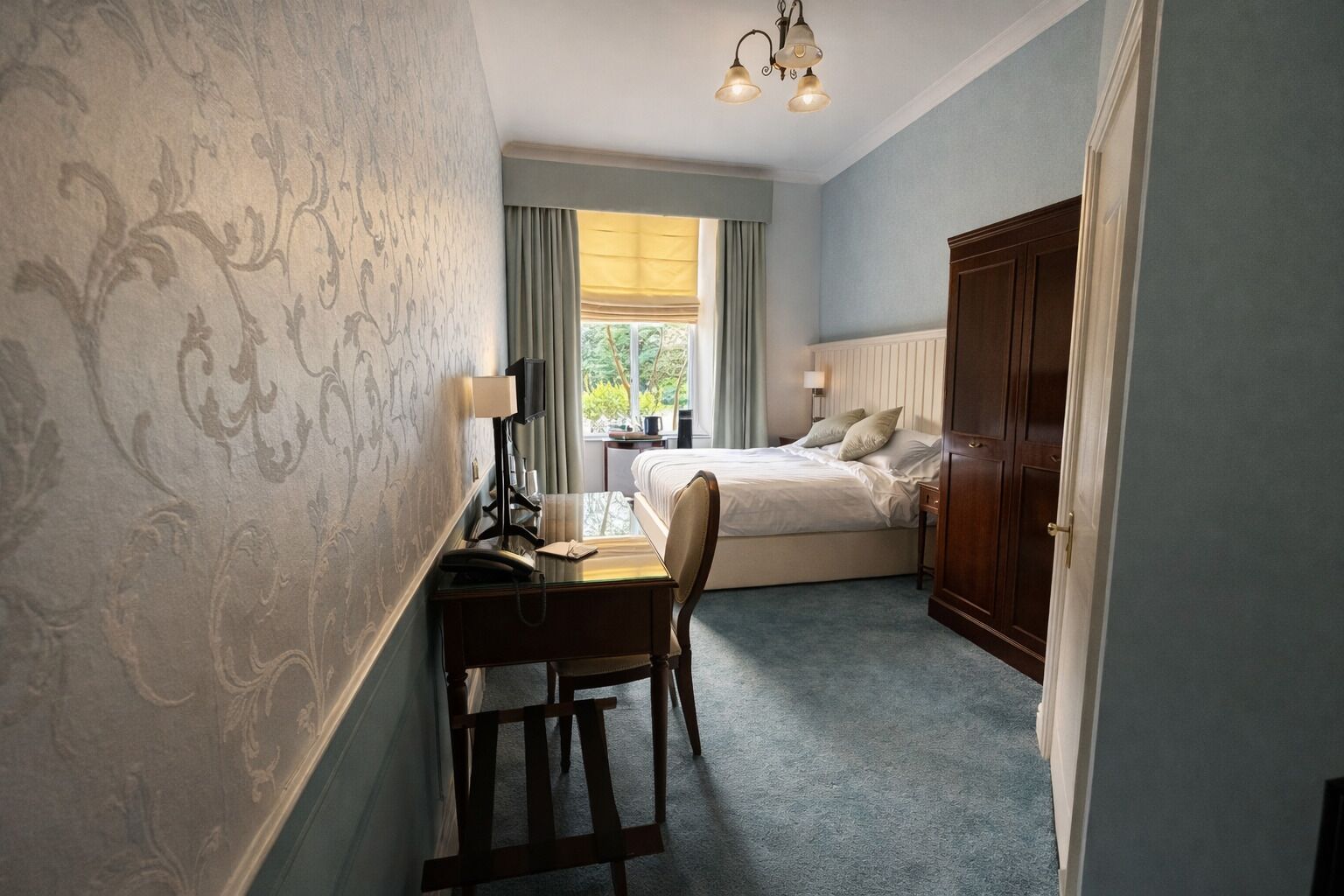Quarto de hotel com cama, escrivaninha com cadeira, guarda-roupa e janela com cortinas.