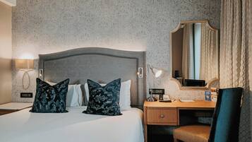 Quarto de hotel com cama, abajures, escrivaninha com cadeira, espelho e papel de parede estampado.