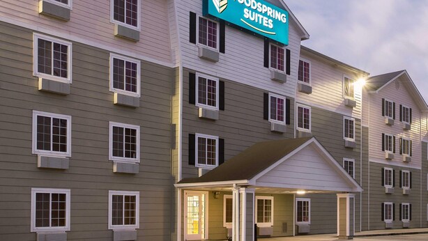 Um hotel de vários andares com uma placa indicando "Woodspring Suites". Possui entrada coberta com teto, várias janelas e aparelhos de ar condicionado.
