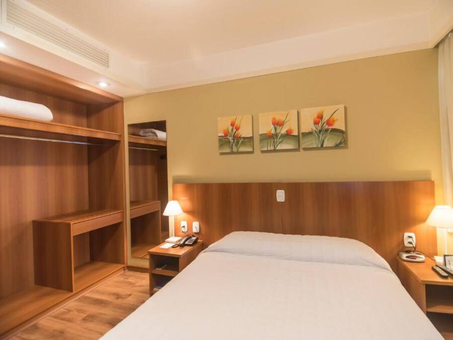 Quarto de hotel com uma cama grande, guarda-roupa de madeira, mesas de cabeceira com luminárias e uma obra de arte floral na parede.