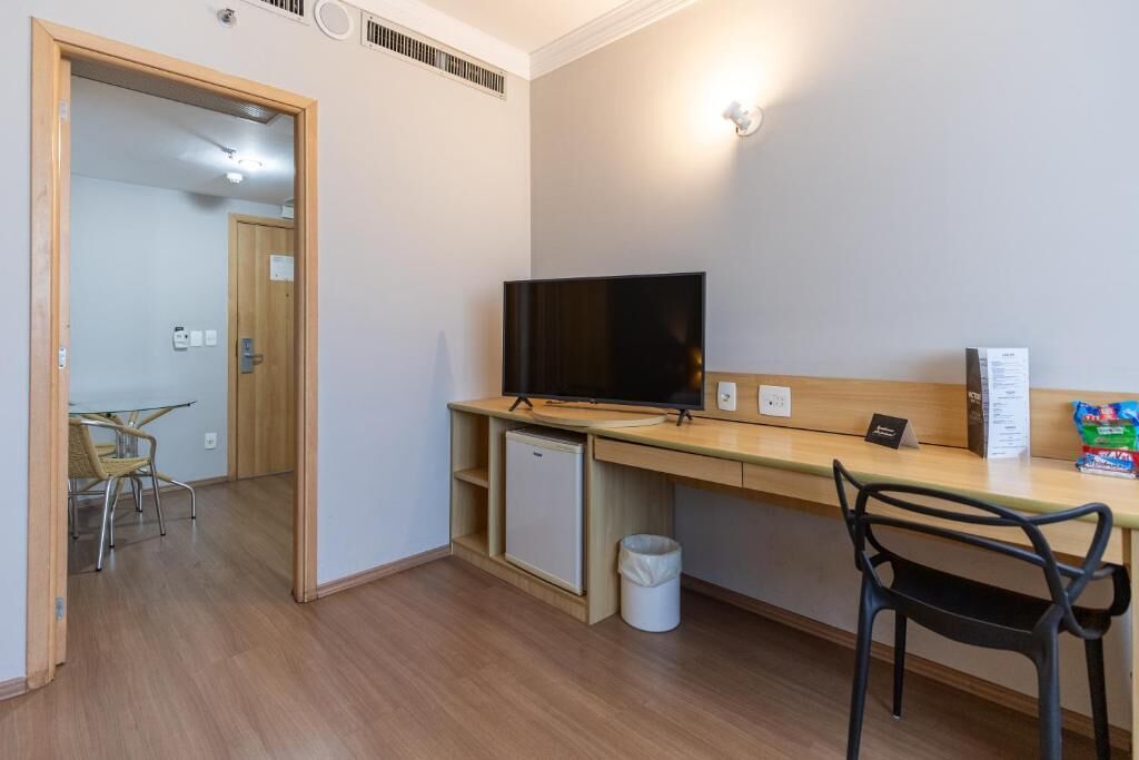 Quarto compacto com uma escrivaninha de madeira, televisão, pequeno frigorífico e um cesto de lixo.
