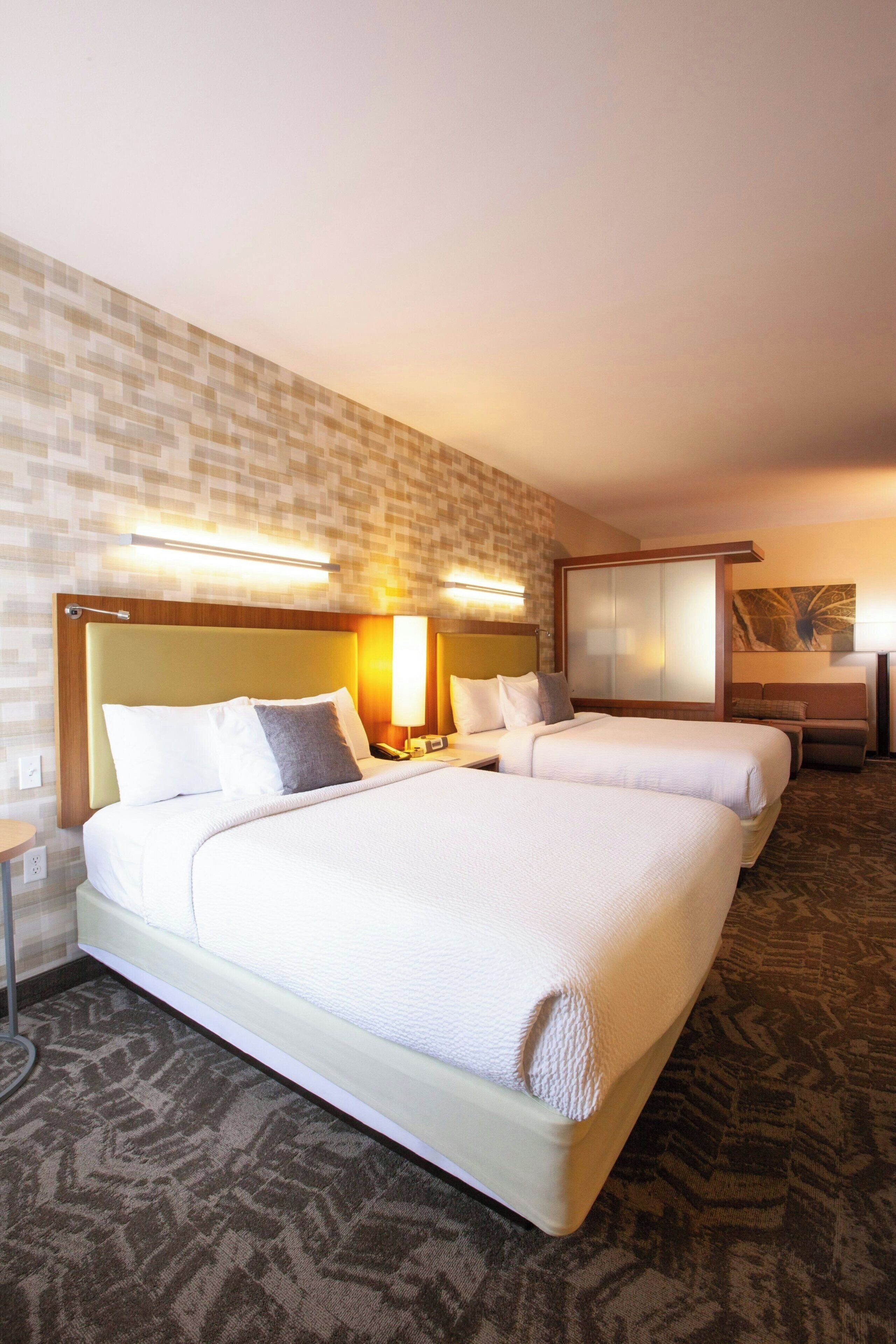 Quarto de hotel com duas camas, cabeceira de madeira, mesa de cabeceira com luminária, obra de arte na parede e tapete estampado.
