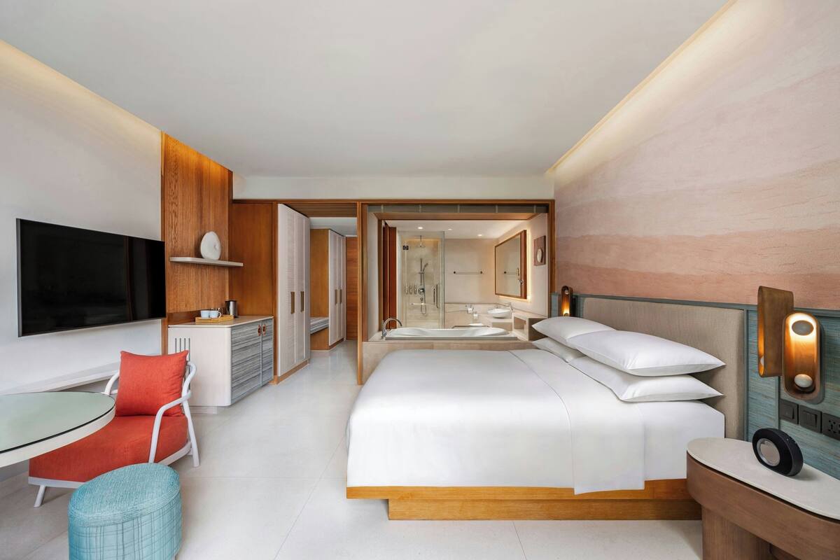 Quarto de hotel moderno com uma cama grande, uma escrivaninha com cadeira, televisão e banheiro visível por uma porta aberta.