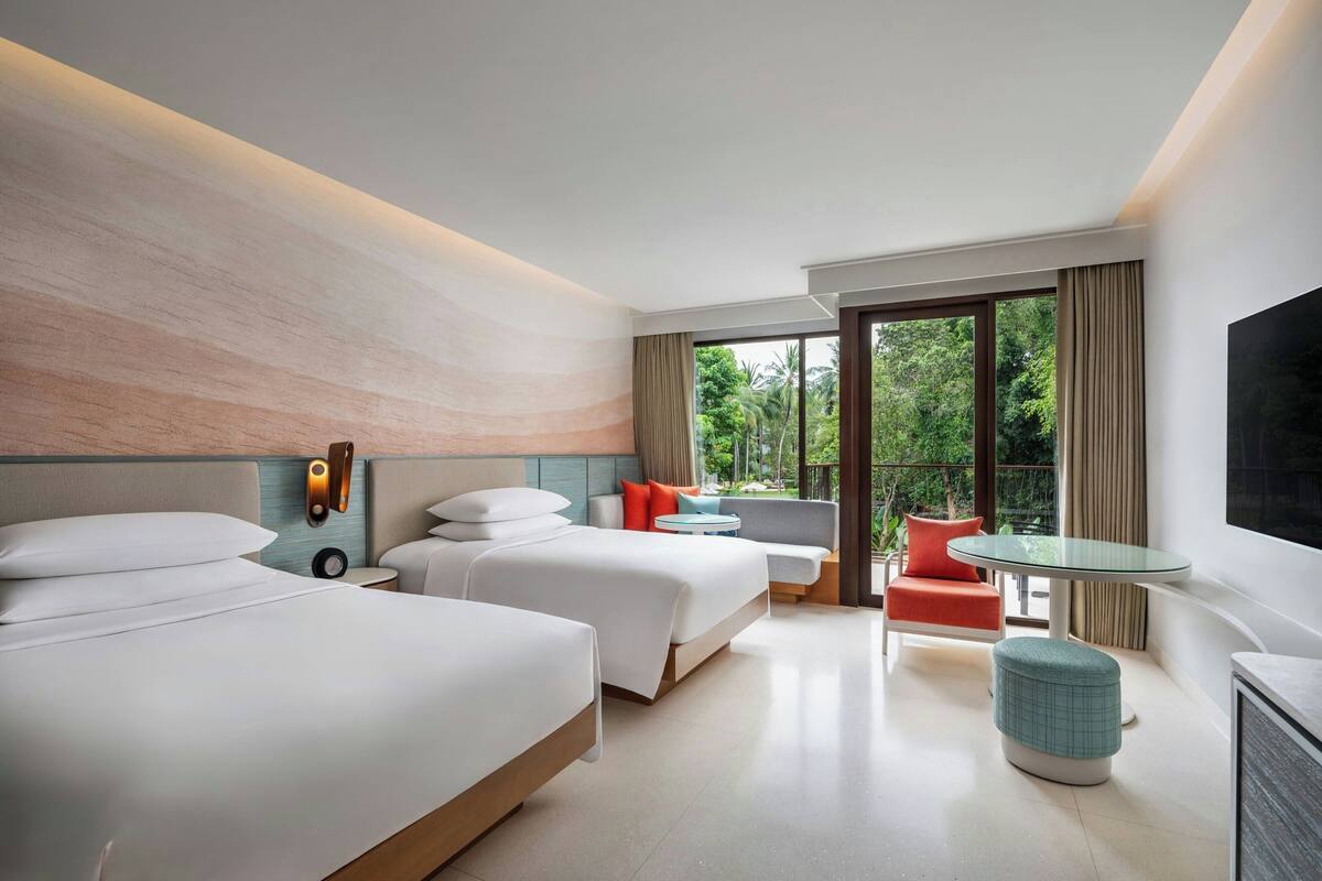 Quarto de hotel moderno com duas camas, uma mesa de vidro, um sofá e uma televisão.