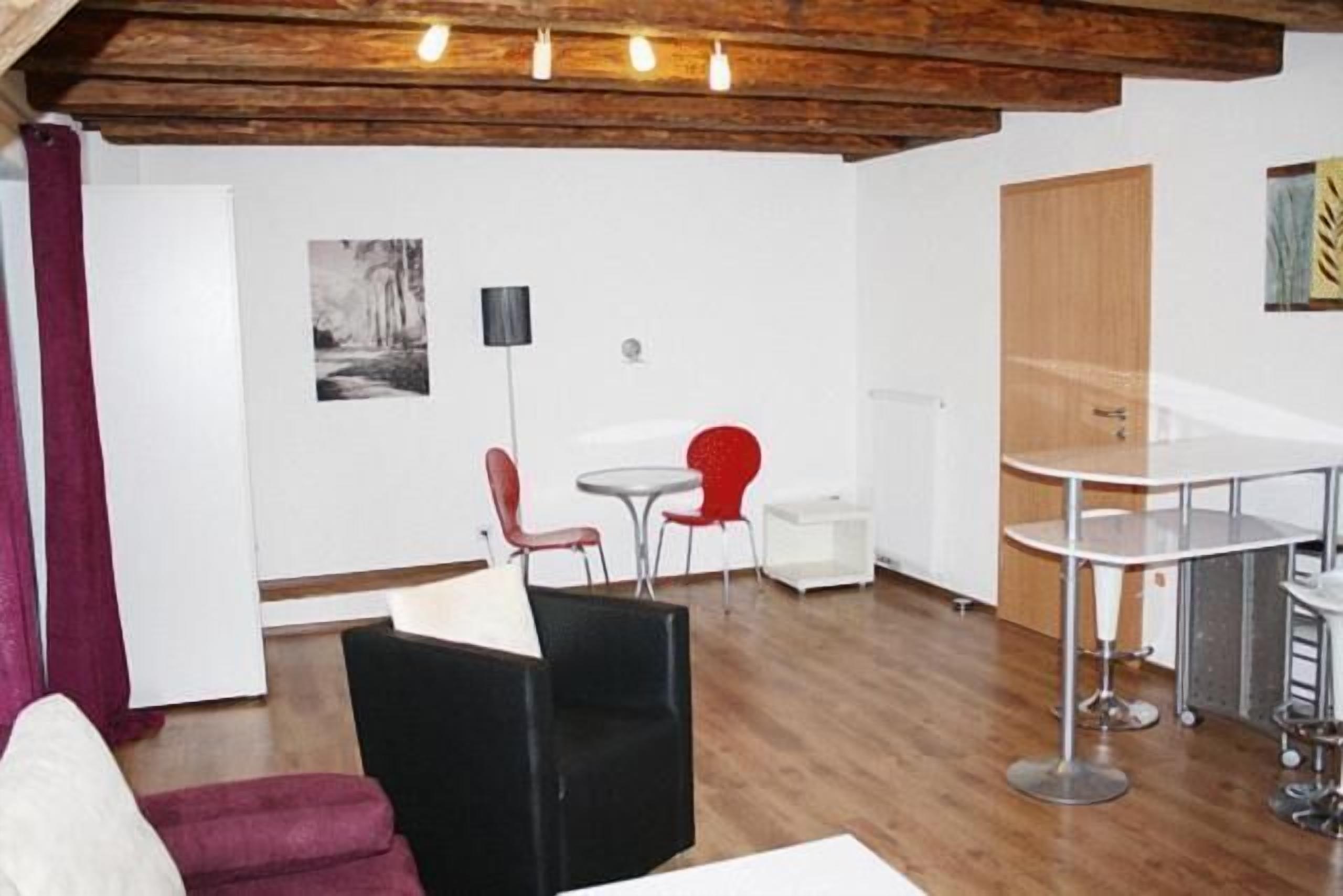 Quarto moderno com teto de madeira, sofá preto, cadeira vermelha, mesa branca e um pequeno armário branco.