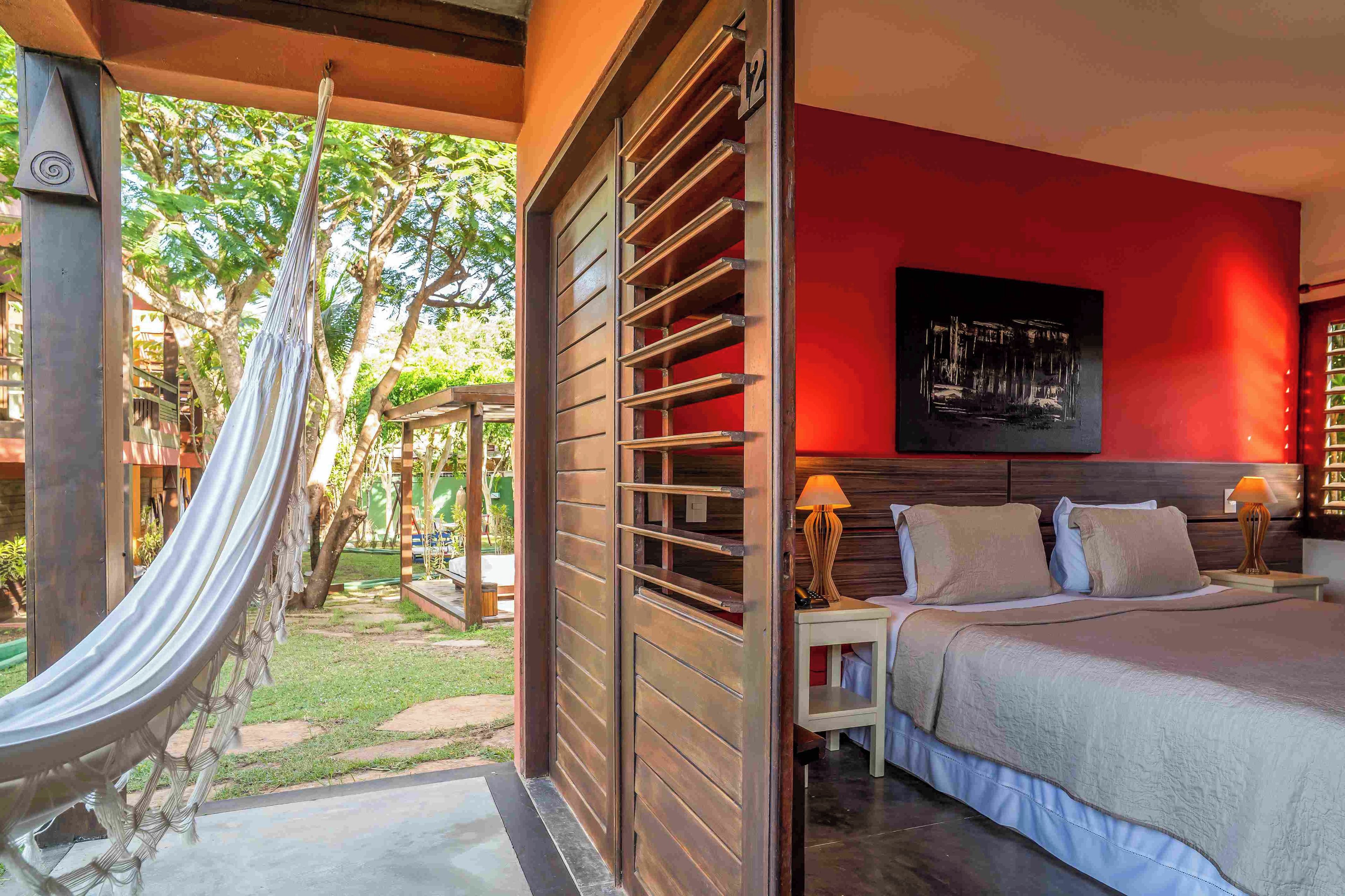 Quarto com cama, luminárias de cabeceira, uma rede e vista para o exterior.