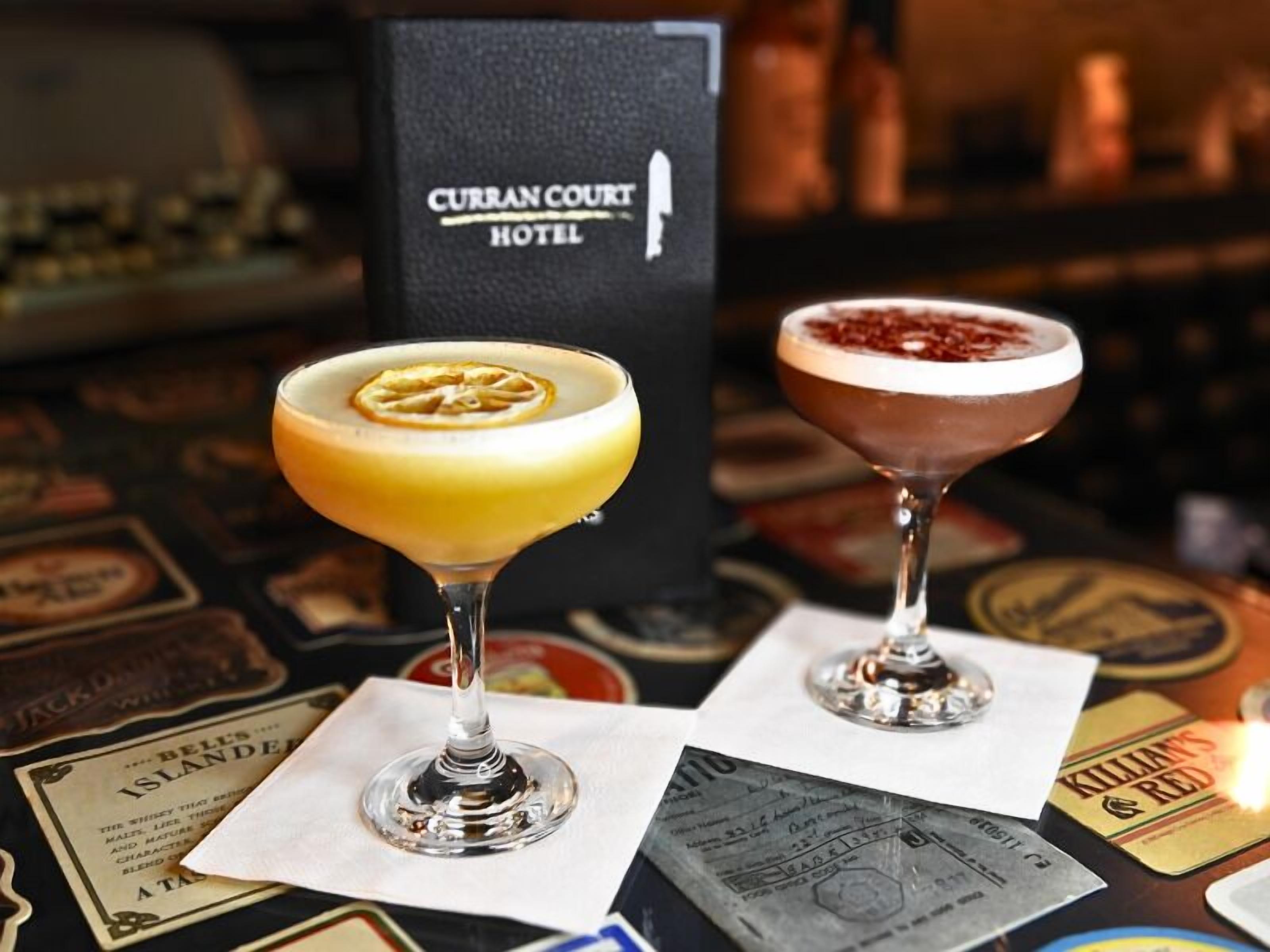 Dois cocktails sobre um balcão de bar, com uma caixa preta com a inscrição "CURRAN COURT HOTEL" ao fundo.