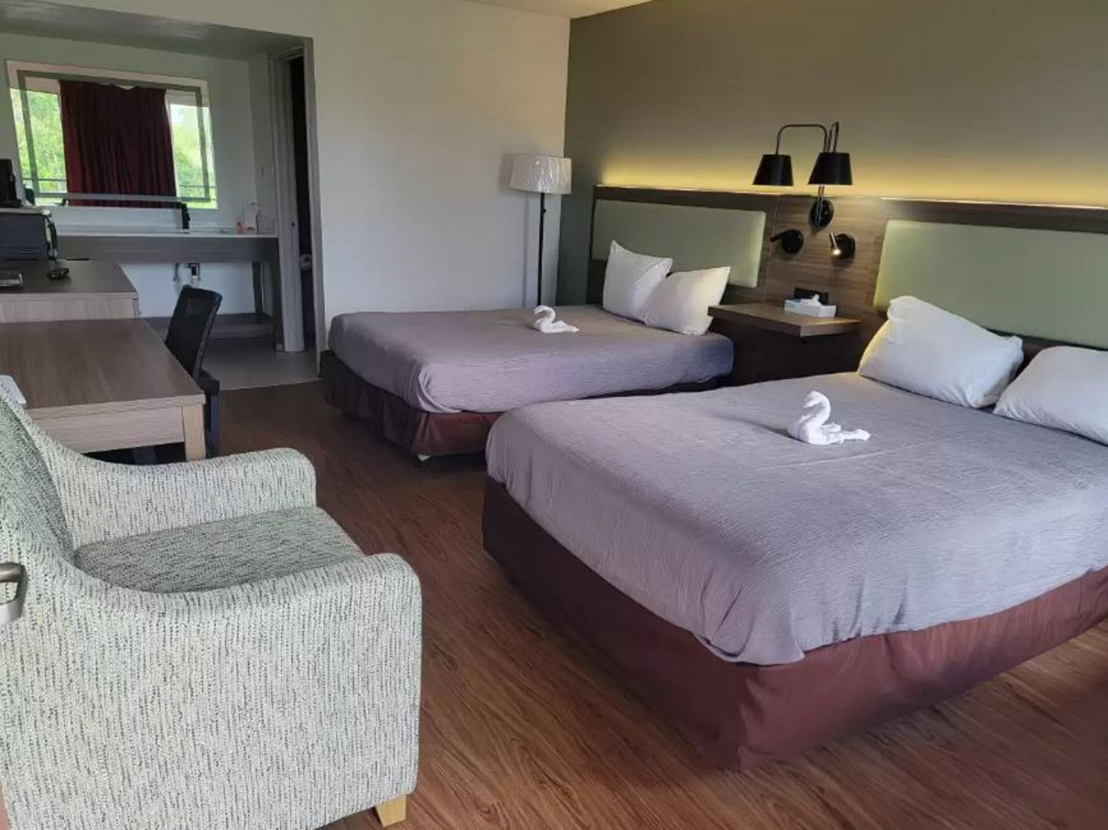 Quarto de hotel com duas camas, uma escrivaninha, uma cadeira e uma luminária.