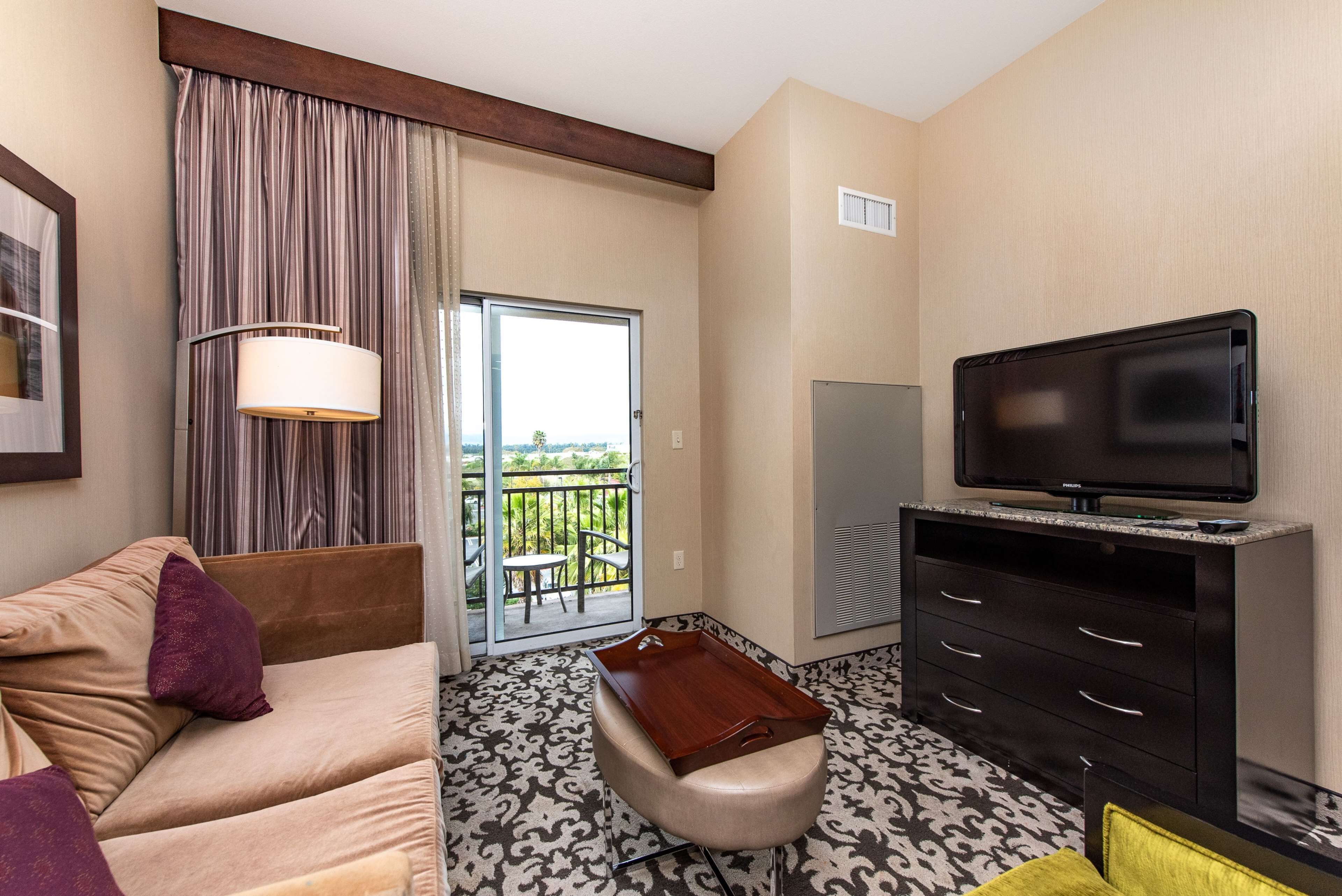 Quarto de hotel com sofá, um pequeno banquinho, televisão e porta de correr para uma varanda.