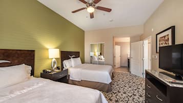 Quarto de hotel com duas camas, ventoinha no teto, televisão, escrivaninha com luminária e banheiro visível por uma porta aberta.