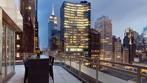 Terraço no telhado com mesa e cadeiras de vidro, com vista para um horizonte urbano de edifícios altos.