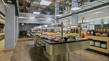 Uma área de buffet com diversos tipos de comida, um espaço de bebidas de serviço e um espaço de refeições com mesas e cadeiras.