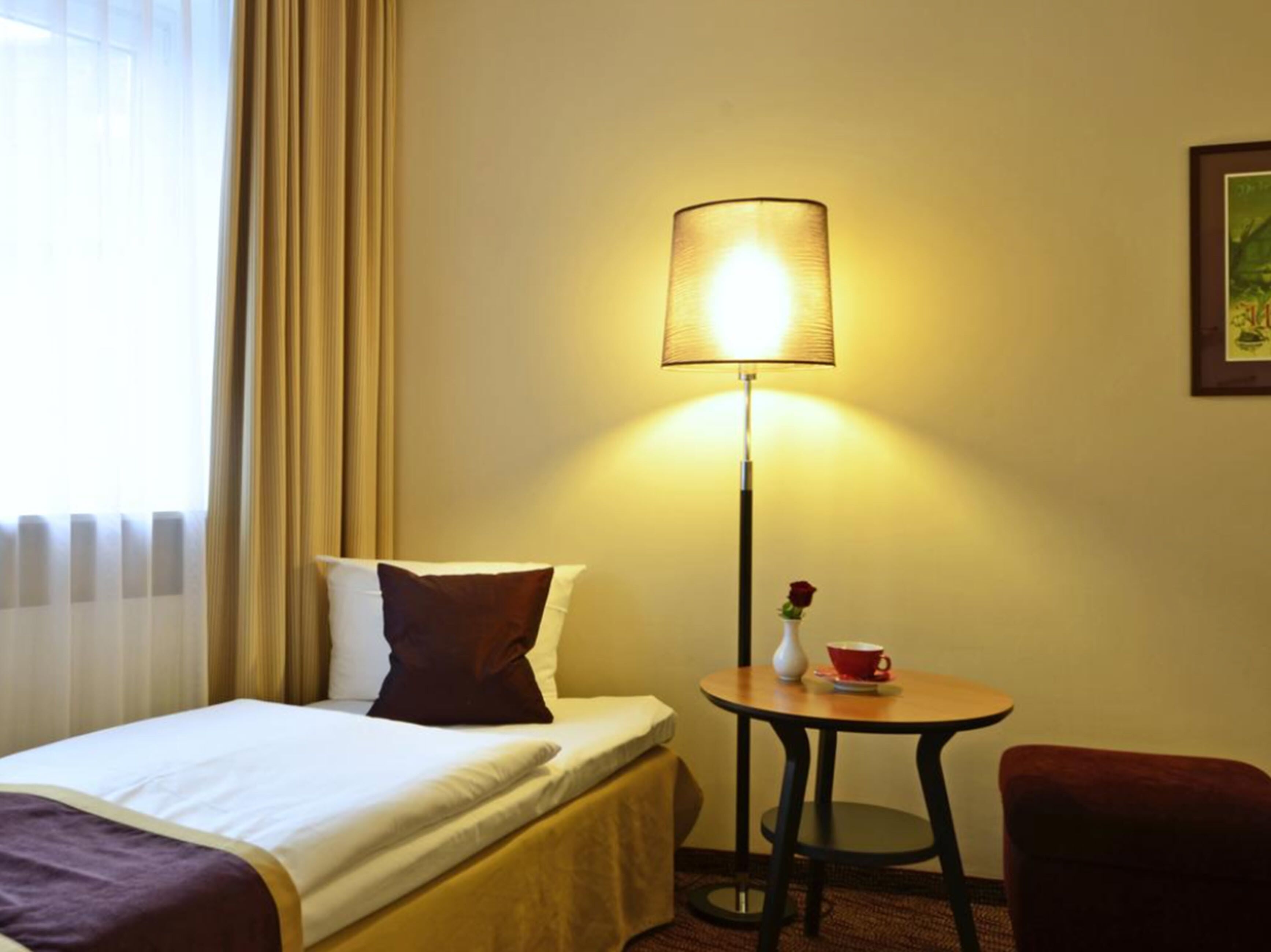 Quarto de hotel com uma cama, uma mesa de cabeceira com abajur, uma mesinha com um vaso e uma xícara, e uma cadeira.
