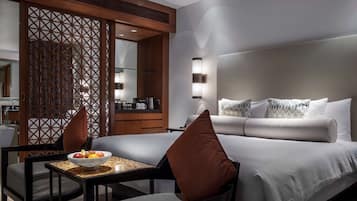 Quarto de hotel moderno com uma cama grande, uma mesa de jantar com uma tigela de frutas e uma área de estar separada.