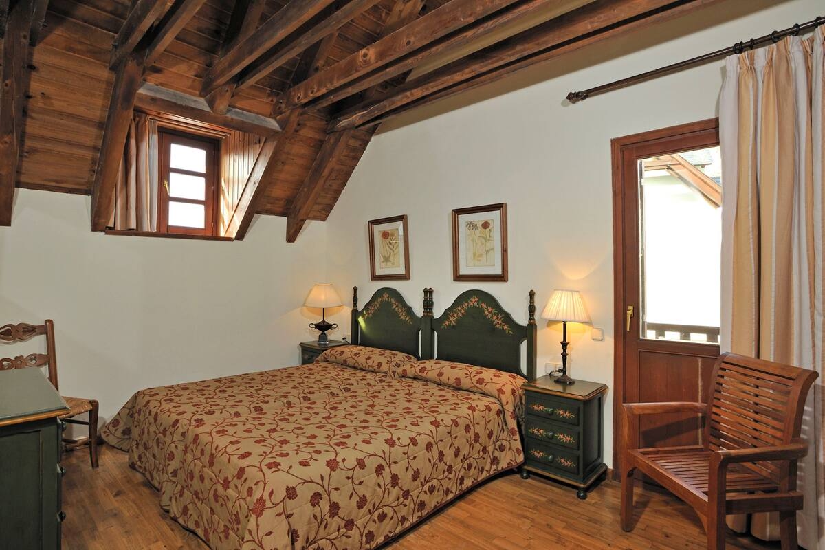 Quarto com teto de madeira, cama com roupa de cama florida, duas mesas de cabeceira com abajures, uma cadeira e janela com cortinas.