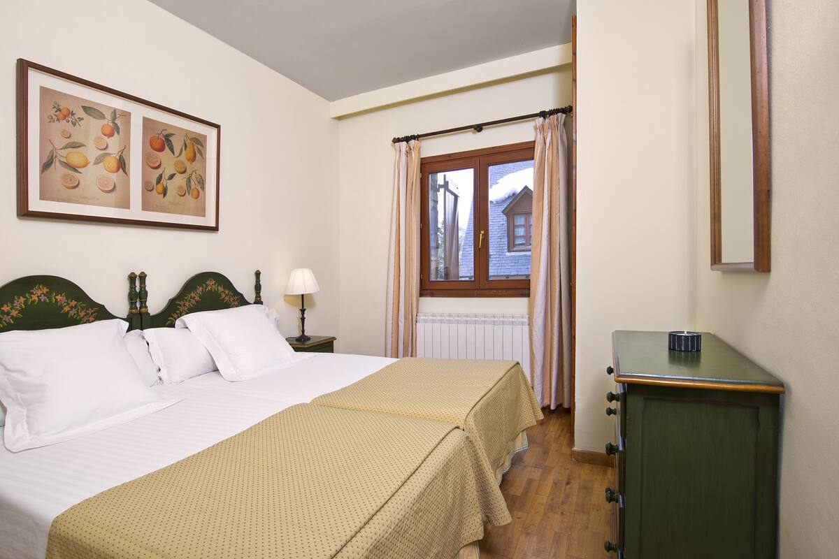 Quarto com cama, duas mesas de cabeceira, janela com cortinas e uma impressão botânica emoldurada na parede.
