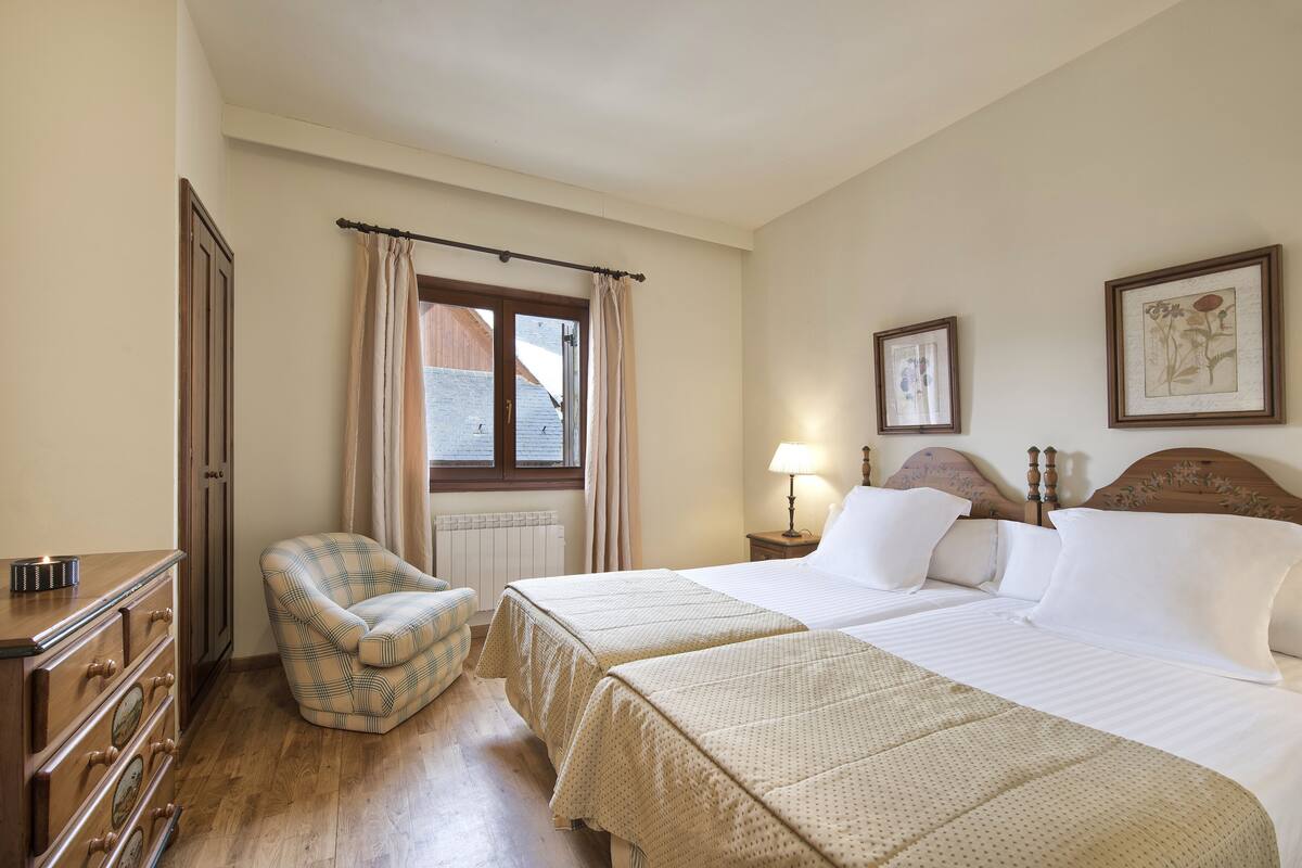 Quarto de hotel com duas camas, cômoda de madeira, uma cadeira, janela com cortinas e quadros nas paredes.