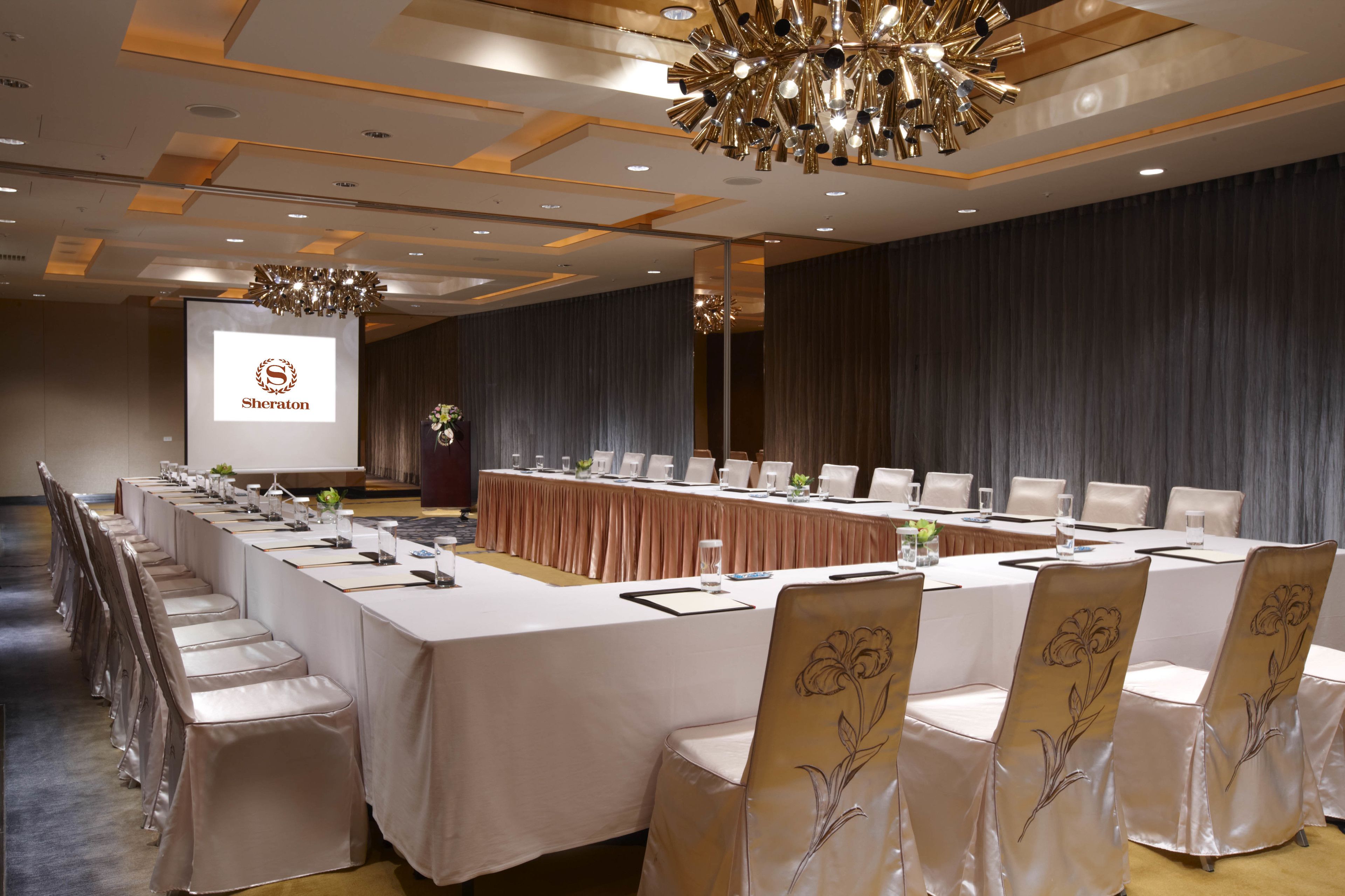 Uma sala de conferências com uma mesa comprida, cadeiras e uma tela exibindo o logotipo Sheraton.